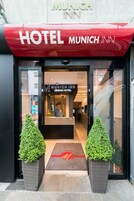 A entrada do Hotel Design Munich Inn, com toldo vermelho e duas plantas em vasos.