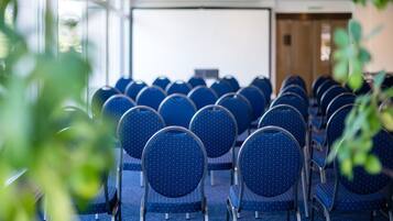 Fileiras de cadeiras azuis com estrutura metálica em uma sala de conferências com grandes janelas e uma tela de projetor.