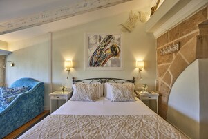 Quarto com uma cama grande, duas mesinhas de cabeceira, uma cadeira, um quadro na parede e uma parede de pedra.