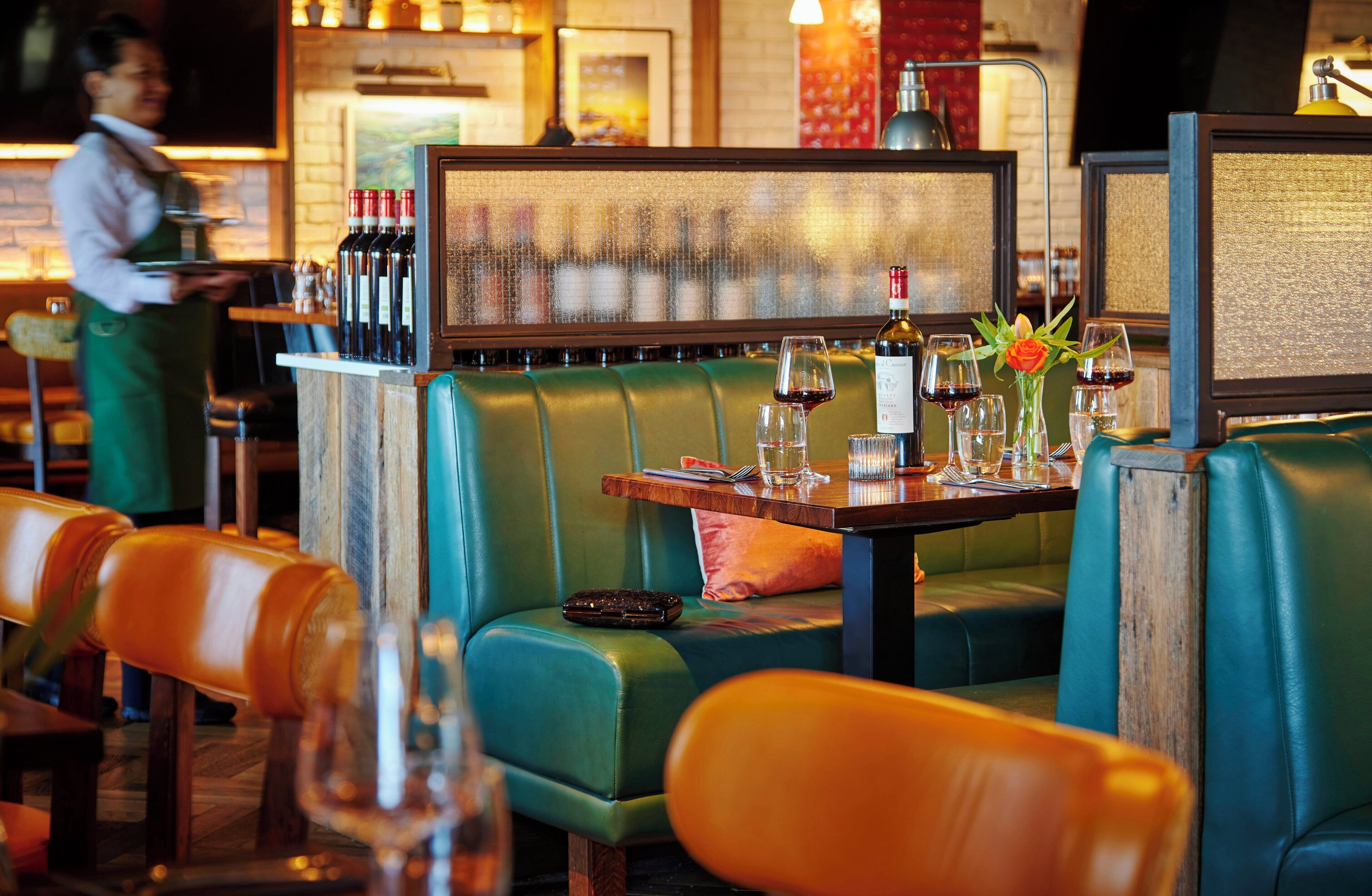 Interior de restaurante com cabines estofadas em verde, área de bar com garrafas e um quadro de cardápio.