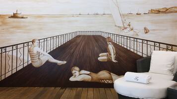 Quarto com piso de madeira, sofá branco e um mural retratando uma paisagem marítima com barcos e um farol.