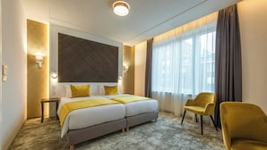 Quarto de hotel com cama, duas mesas de cabeceira, uma televisão grande montada na parede, janela com cortinas e duas cadeiras amarelas.