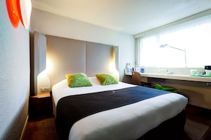 Quarto de hotel com cama, luminárias de cabeceira, escrivaninha com cadeira, janela e mesa de cabeceira.
