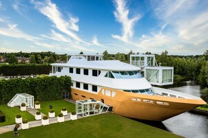 Uma casa-barco moderna com um amplo deck, áreas envidraçadas e um jardim com arbustos bem cuidados.