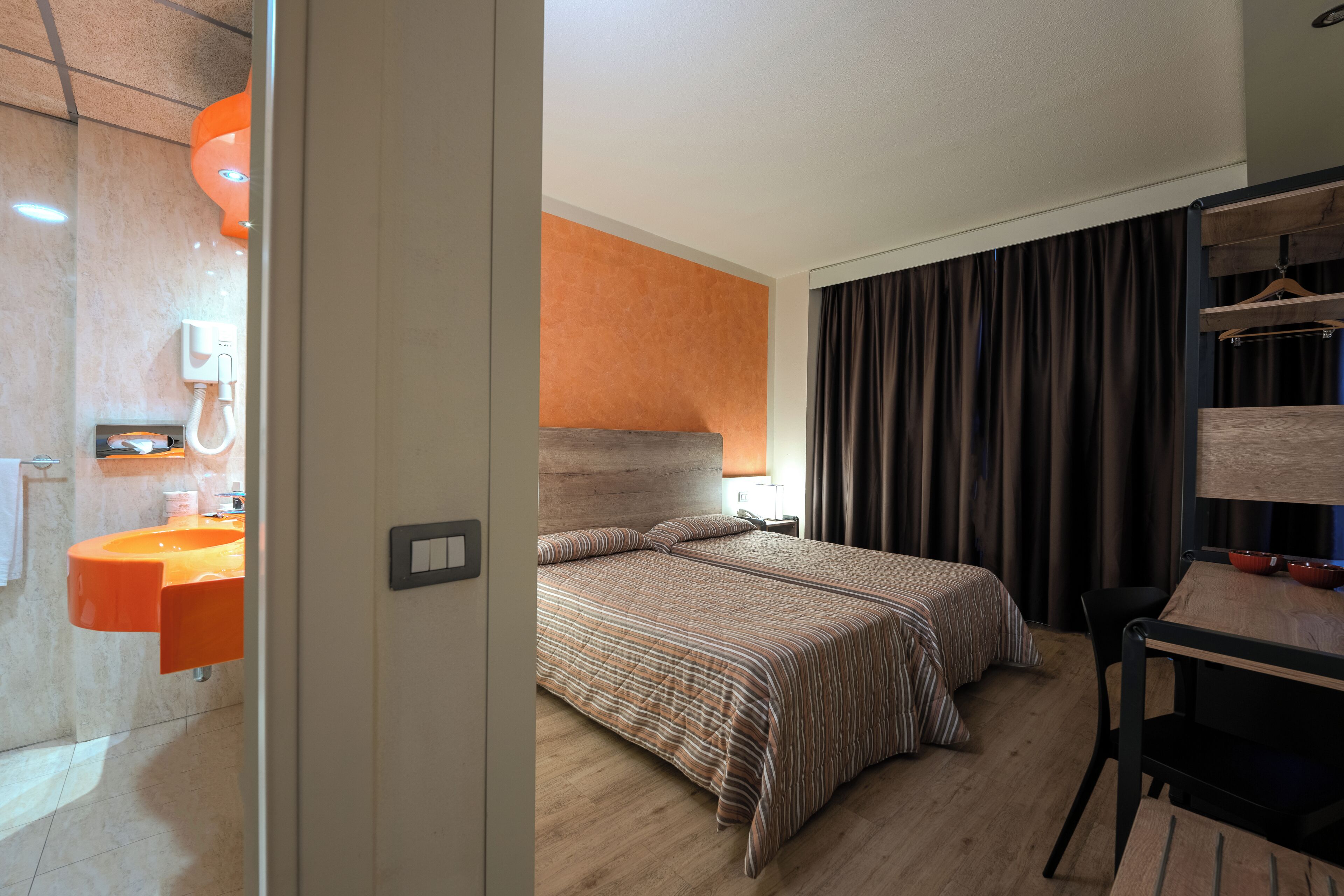 Quarto de hotel moderno com uma cama grande, cabeceira de madeira, mesa de cabeceira com abajur e pia laranja.