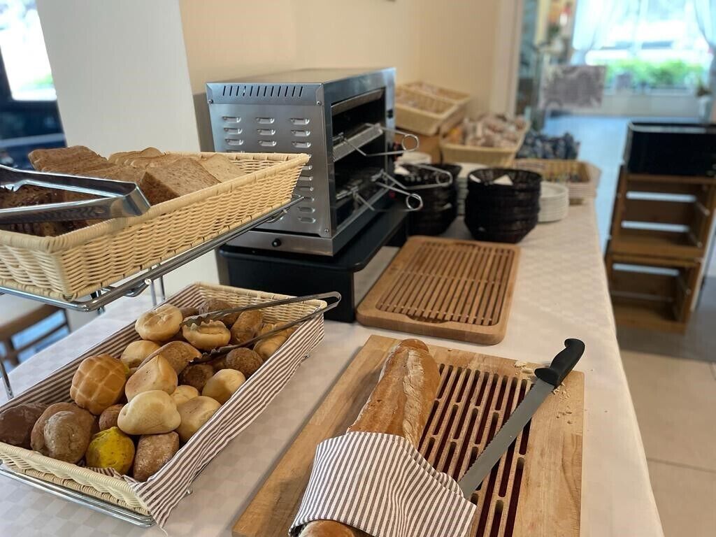 Disposição de buffet com diversos pães e pastéis, um fatiador de pão e cestas.