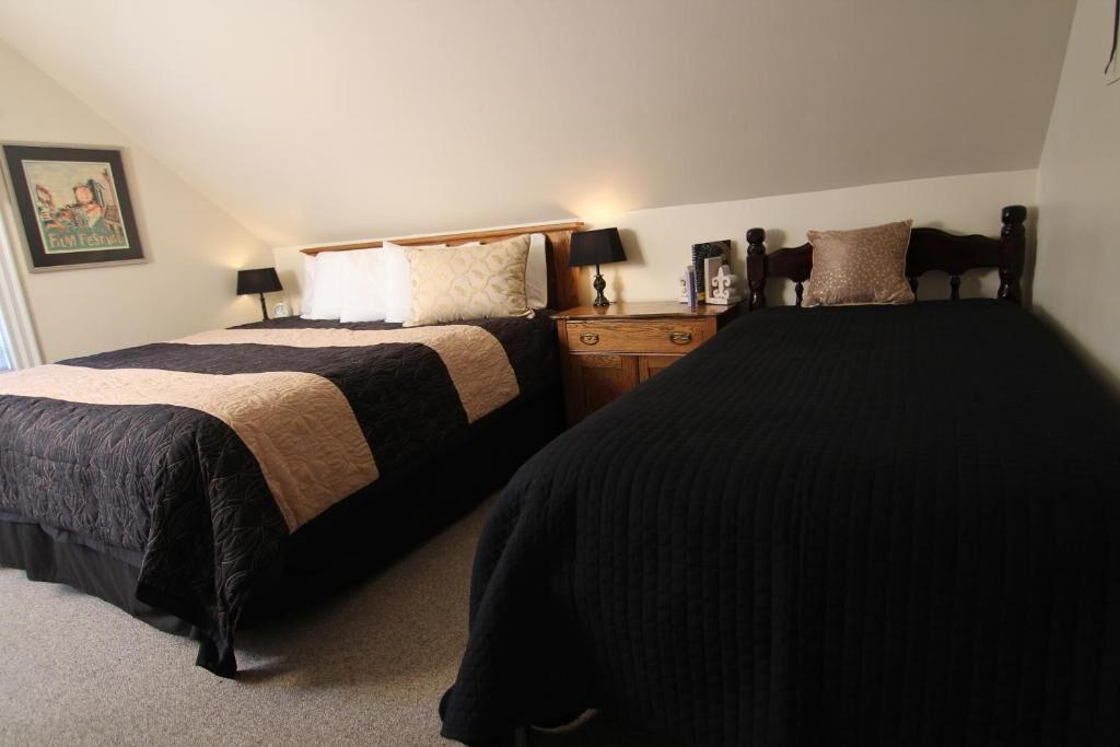Quarto com duas camas, cada uma com um cobreleiro escuro e um cobertor claro. Apresenta uma mesa de cabeceira com uma luminária e uma pequena mesa com um livro e um objeto decorativo.