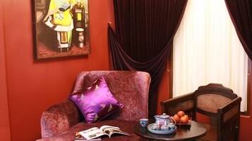 Um quarto com parede vermelha, um sofá vermelho com almofada roxa, uma cadeira de madeira, uma mesa redonda pequena com um bule e laranjas, e um candelabro.