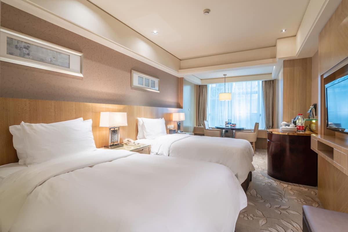 Quarto de hotel com duas camas, uma escrivaninha, uma cadeira, uma televisão e uma janela com cortinas.