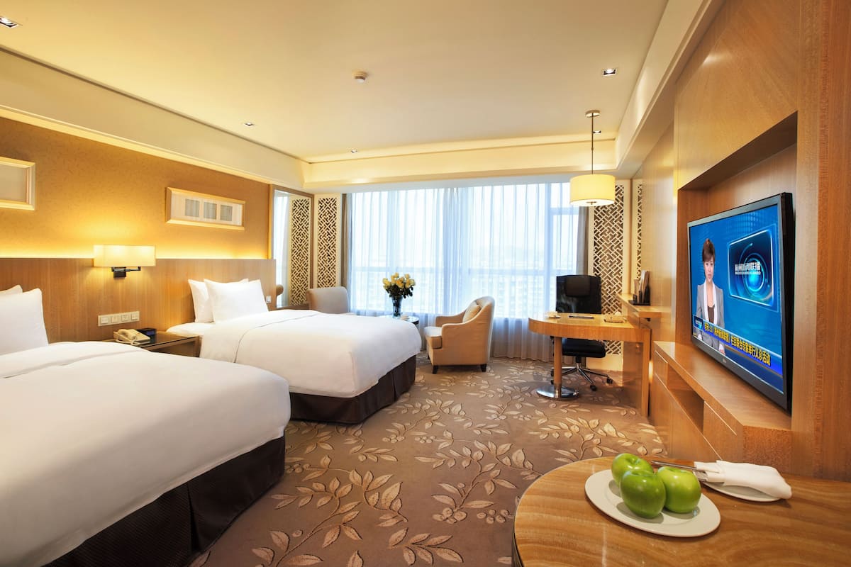 Quarto de hotel com duas camas, uma escrivaninha, uma cadeira, uma TV e uma mesinha com maçãs.