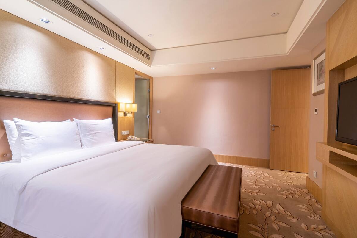 Quarto de hotel com uma cama grande, uma mesa de cabeceira com abajur, uma televisão fixada na parede e uma porta de madeira.