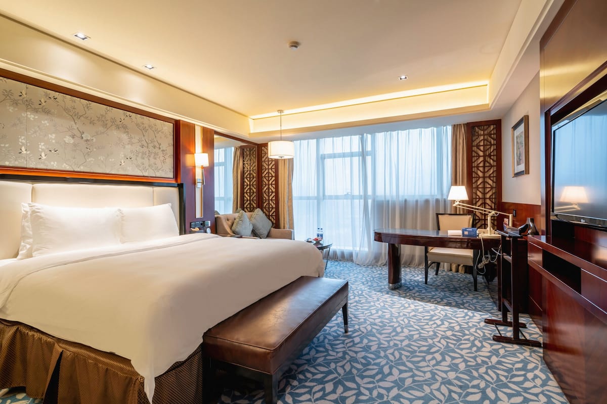 Quarto de hotel com uma cama grande, uma escrivaninha, uma cadeira, um banco e uma televisão.