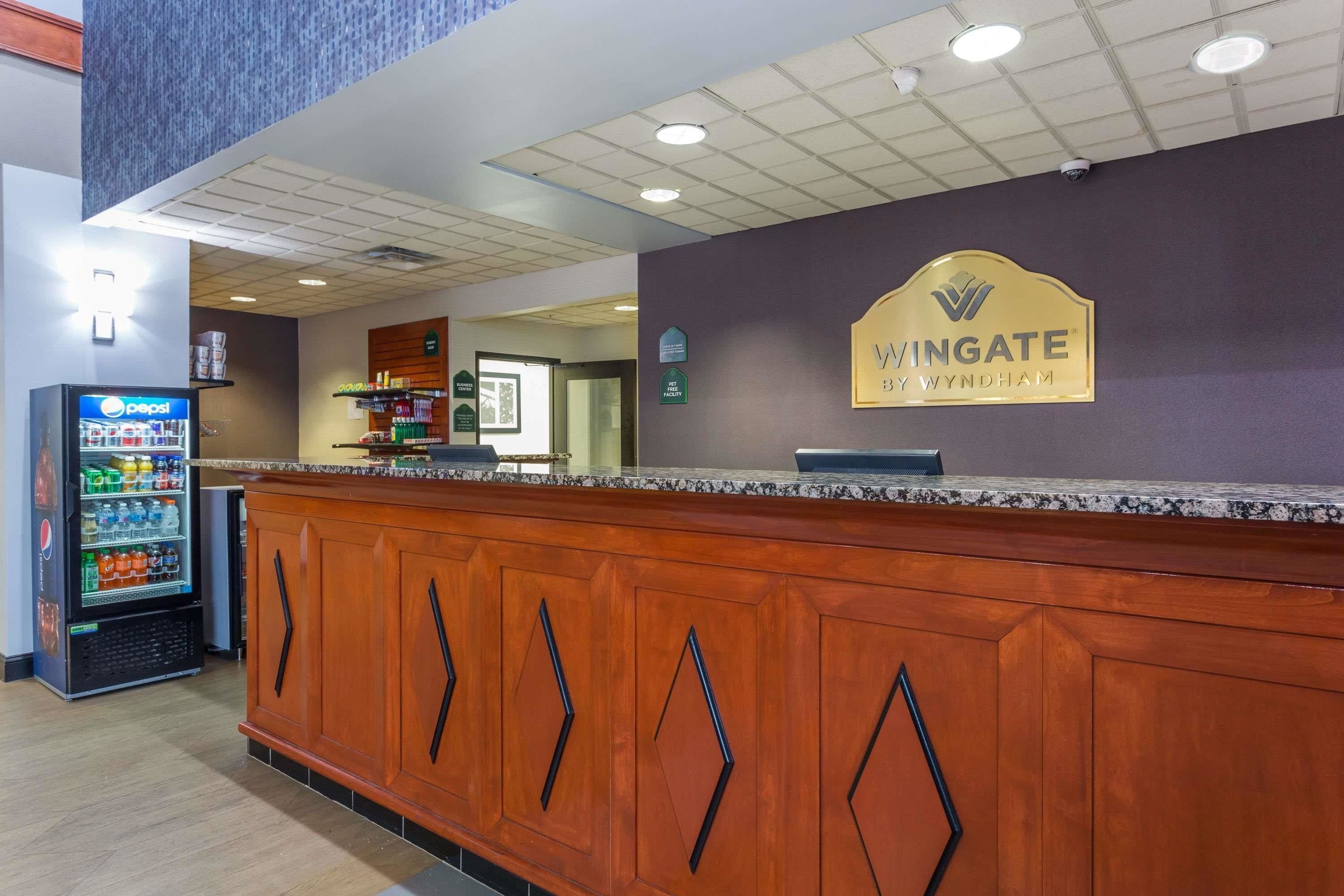 Uma área de recepção com balcão de madeira, refrigerador de Pepsi e uma placa com a inscrição "WINGATE BY WYNDHAM".