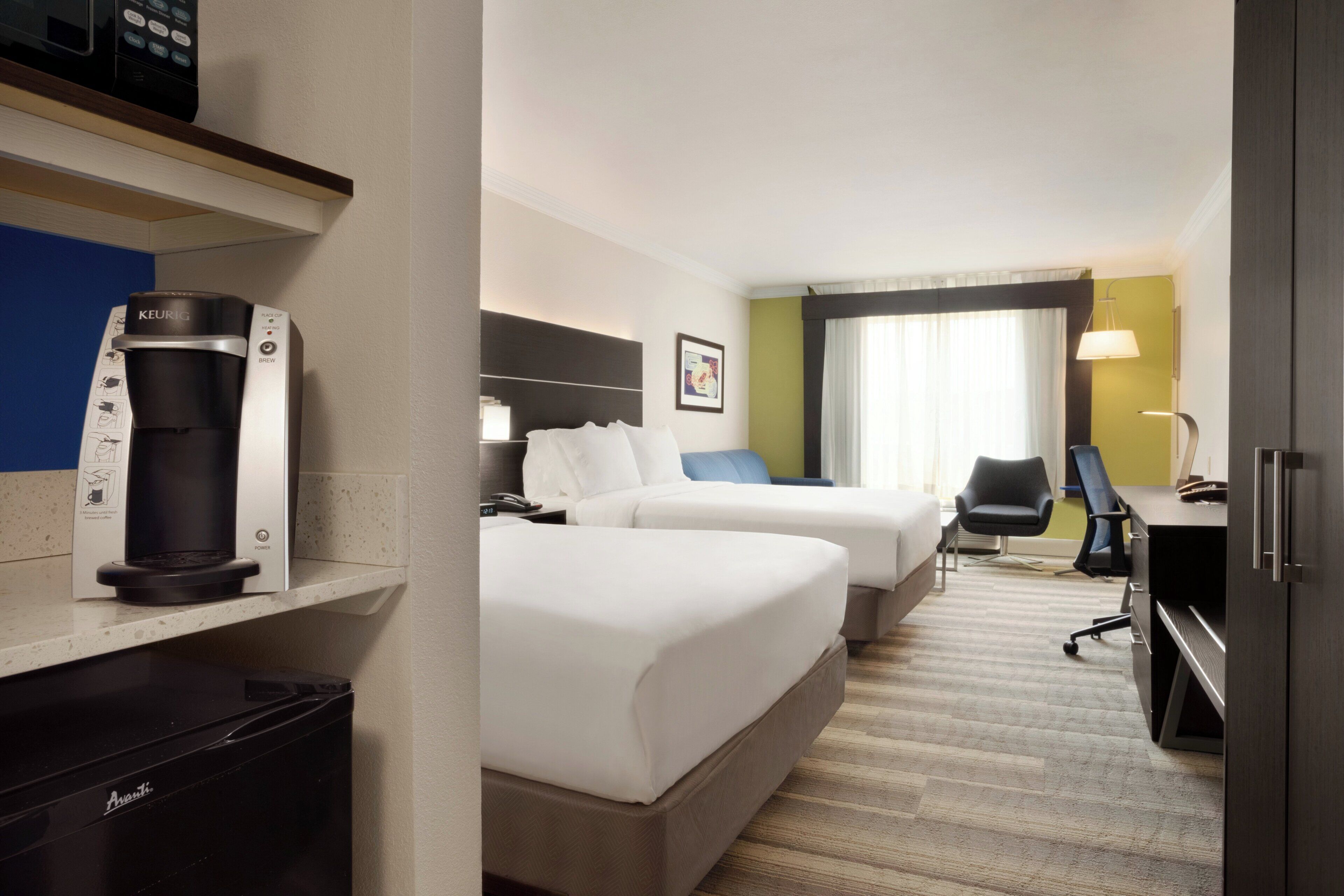 Quarto de hotel com cozinha compacta, uma cama, escrivaninha e uma cadeira.