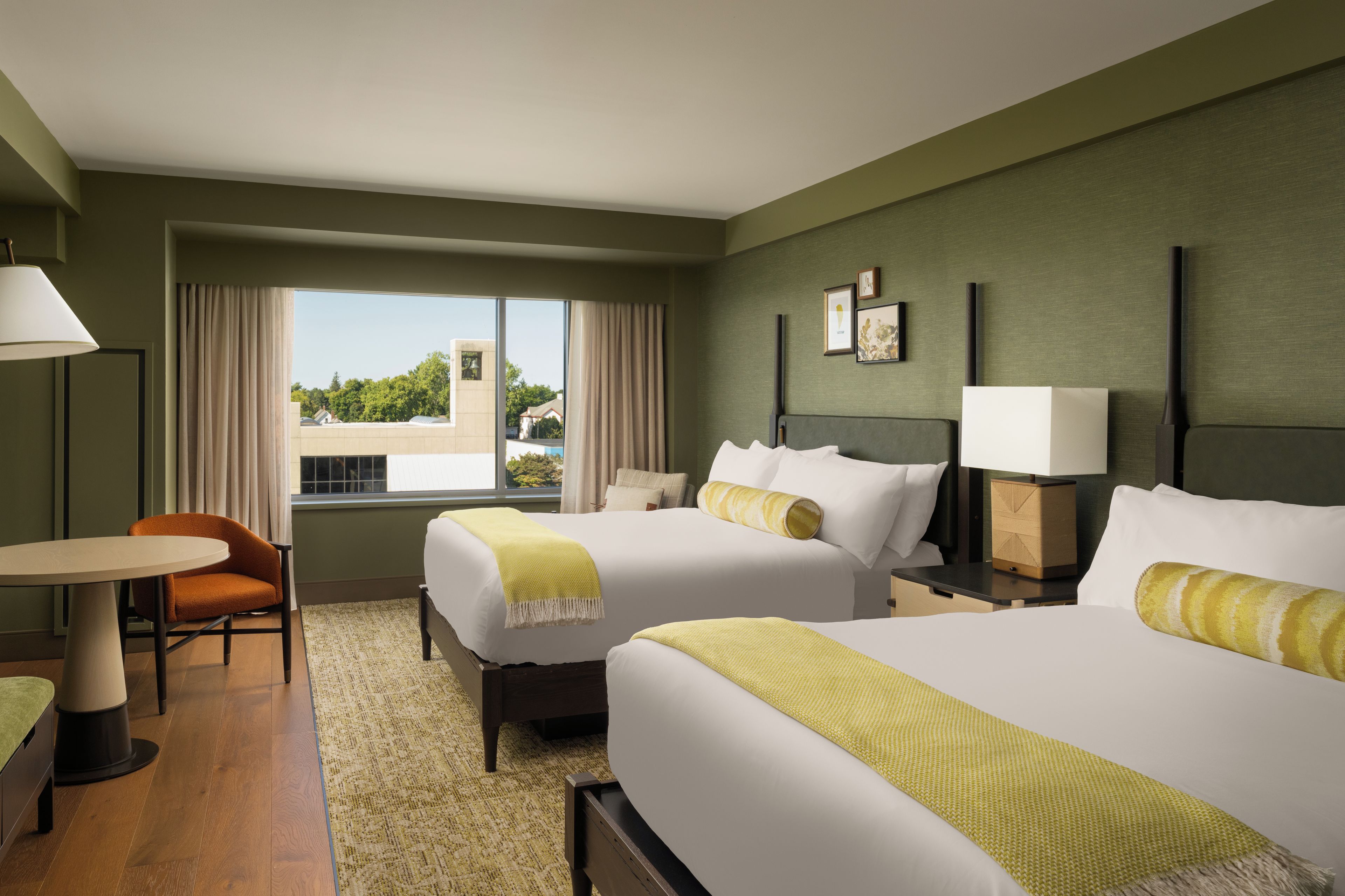 Quarto de hotel com duas camas, uma mesa, uma cadeira e janela com vista para edifícios.