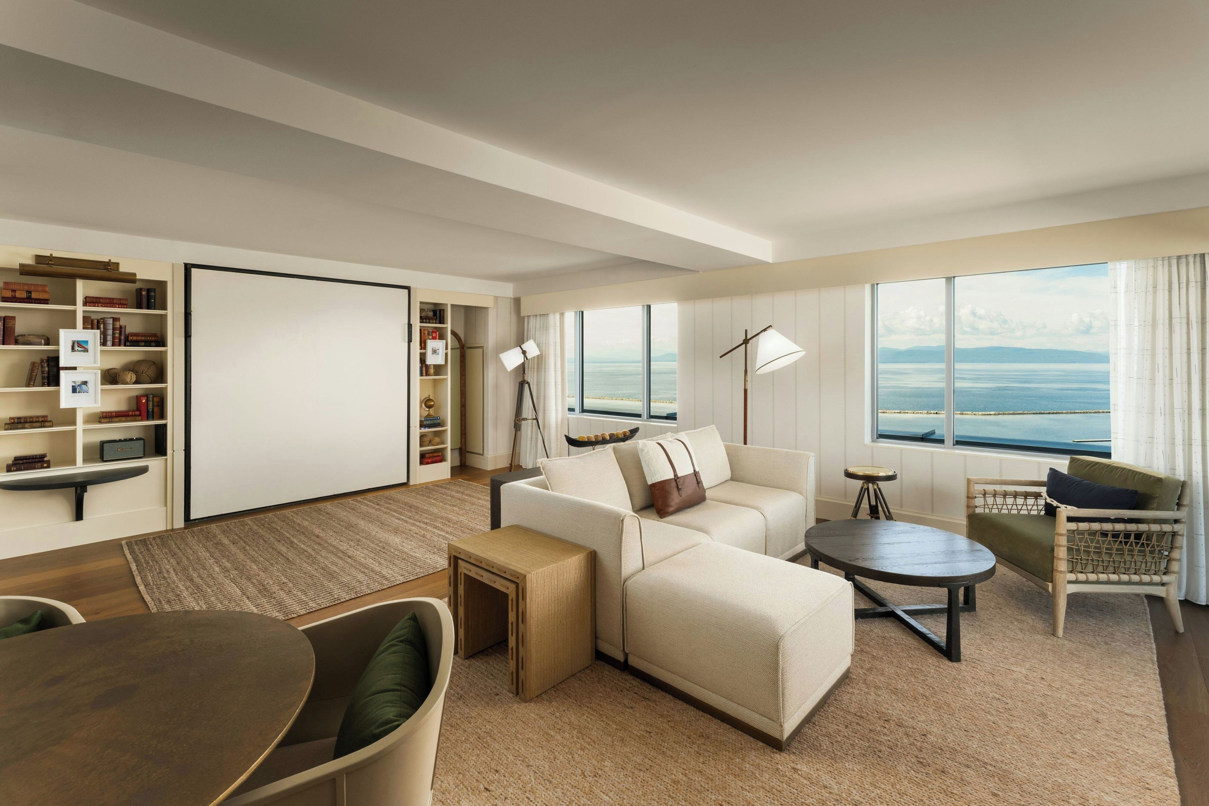 Uma sala de estar moderna com sofá, poltronas, mesa de centro, estante de livros e uma grande janela com vista para o mar.