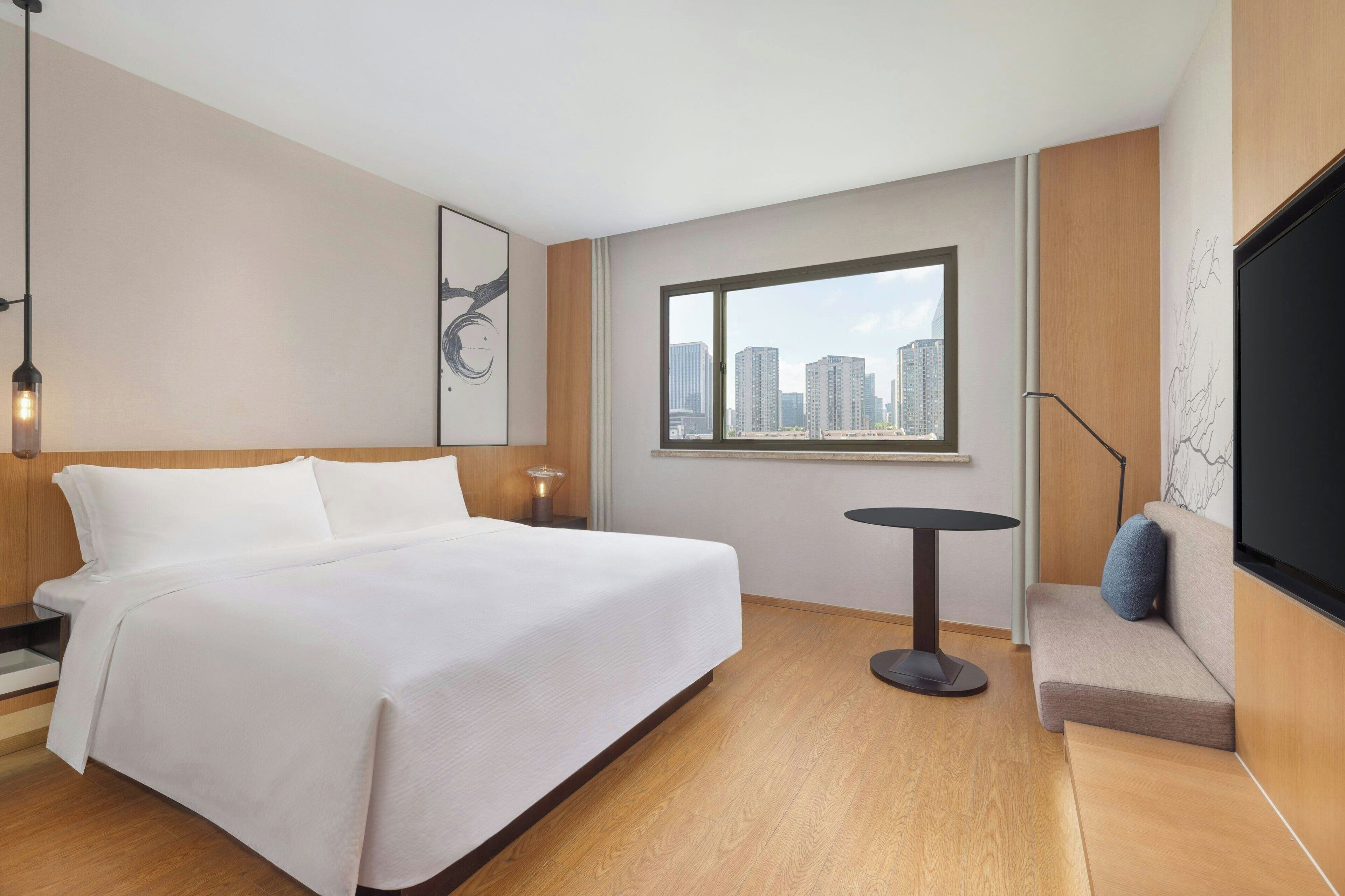 Quarto de hotel moderno com uma cama grande, uma mesa redonda pequena, um sofá, vista para a cidade e uma obra de arte na parede.