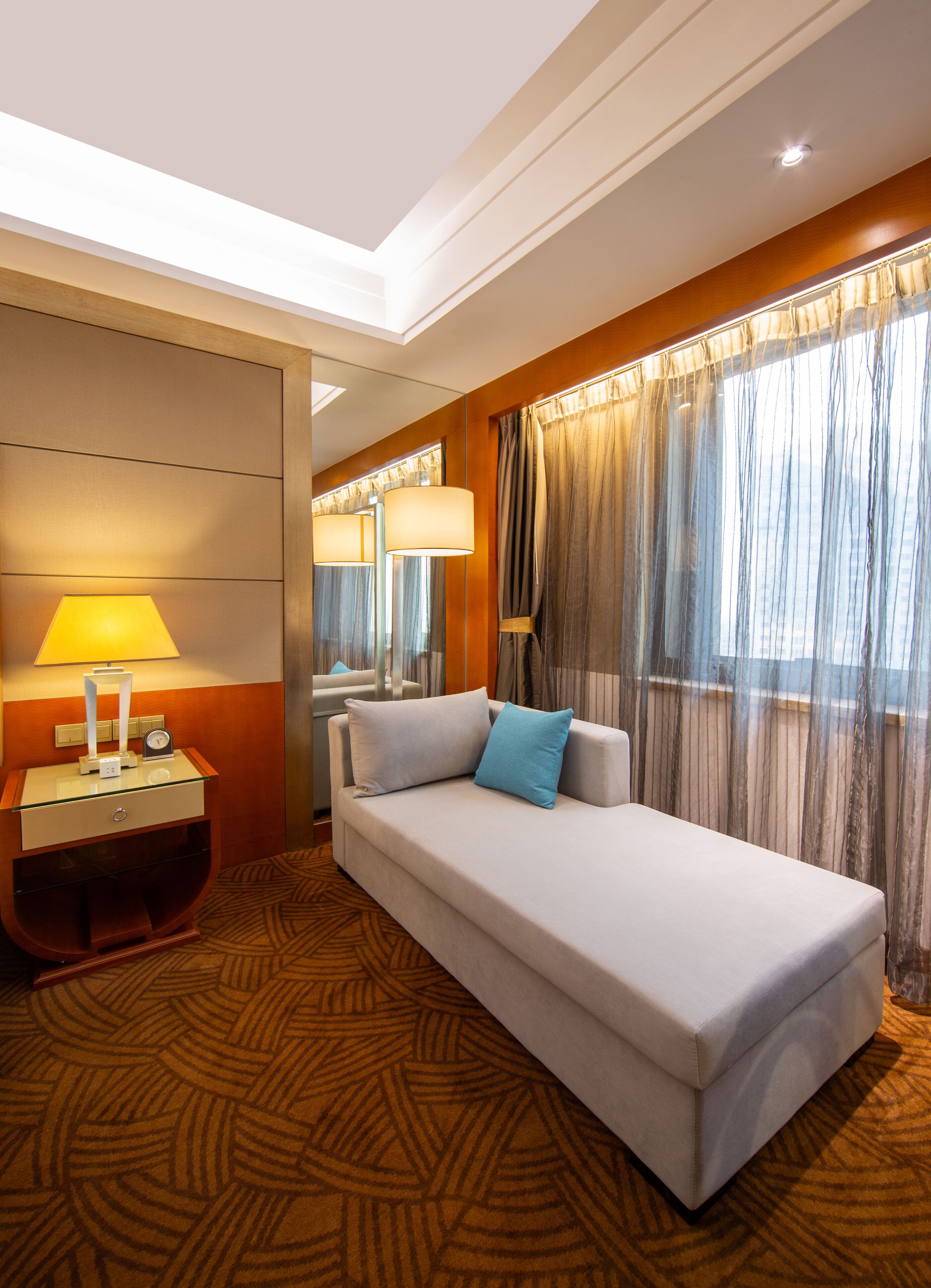Quarto de hotel com uma cama de solteiro, mesa de cabeceira com abajur, espelho e tapete estampado.