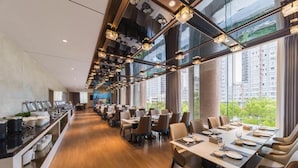 Uma sala de jantar moderna com mesas compridas e cadeiras, um buffet à esquerda e grandes janelas com vista para a cidade.