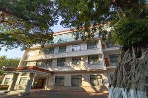 Um edifício de vários andares com entrada central, janelas amplas e uma árvore em primeiro plano.