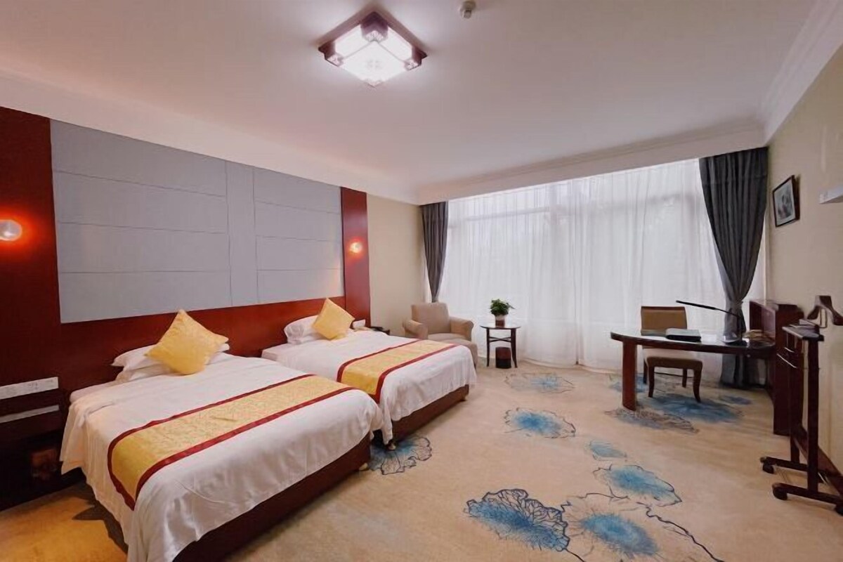 Quarto de hotel com duas camas, uma escrivaninha, uma cadeira, uma mesinha e uma janela grande com cortinas.