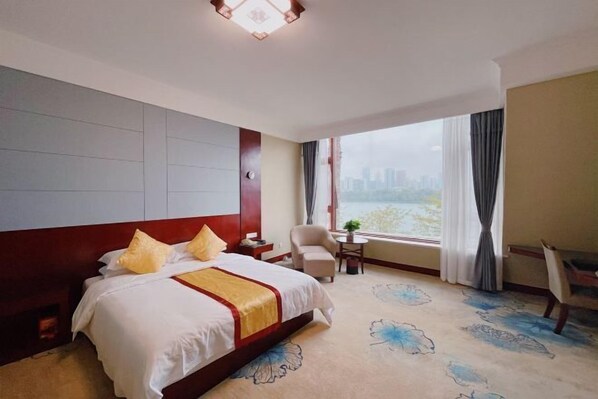 Quarto de hotel com cama, uma cadeira, uma mesinha e vista para a paisagem urbana através de uma janela.