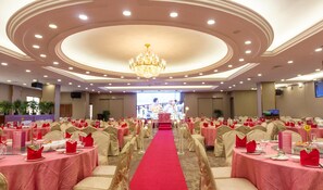 Salão de banquetes com mesas redondas, tapetes vermelhos e um lustre central.