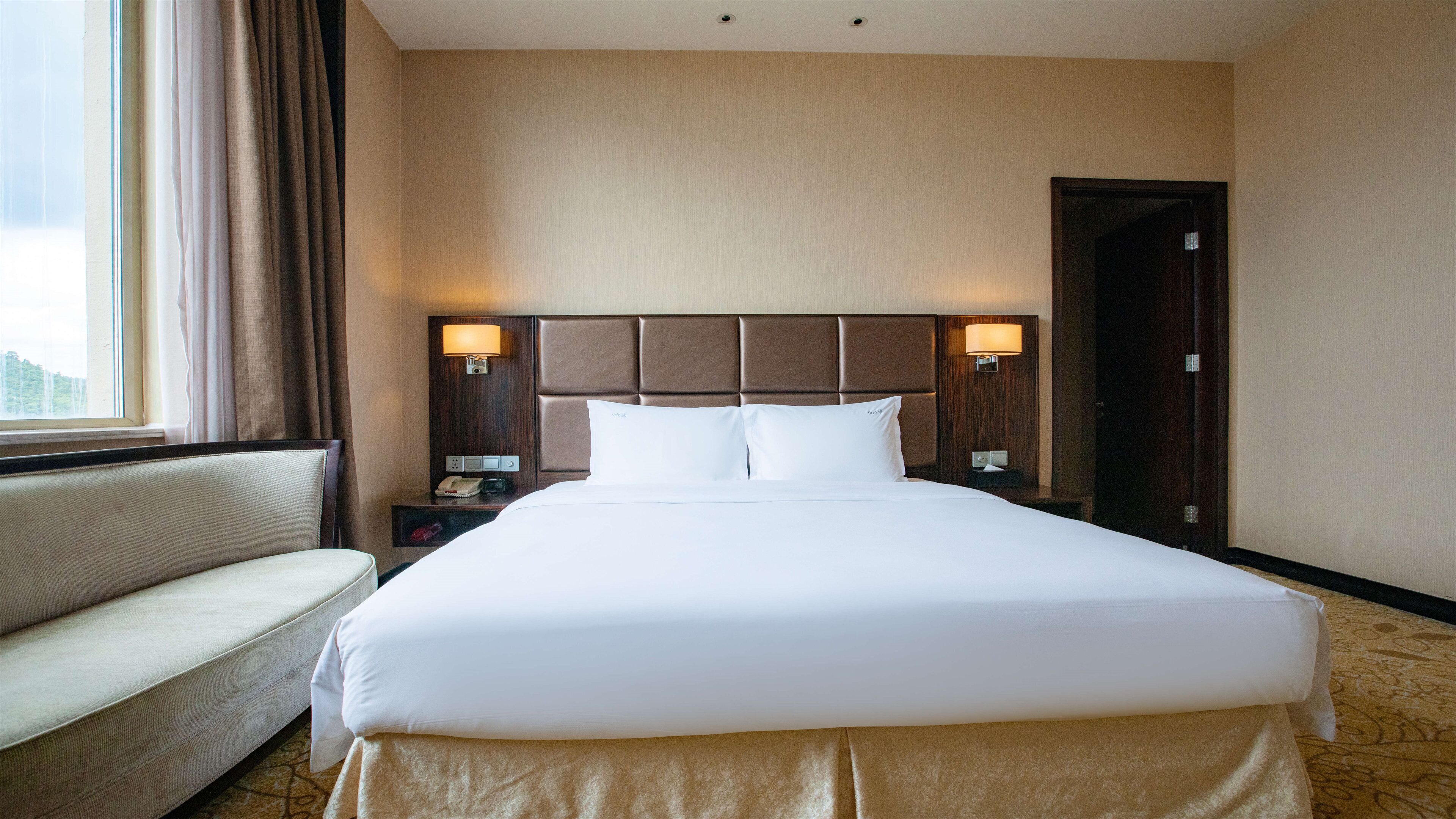 Quarto de hotel com uma cama grande, duas luminárias de cabeceira, uma mesa de apoio, um sofá e uma porta.