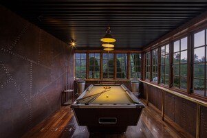 Uma mesa de bilhar em um ambiente com grandes janelas e teto de madeira.