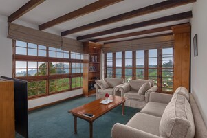 Uma sala de estar ampla com uma grande janela, uma mesa de centro de madeira, um sofá bege e uma estante de livros.