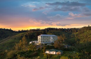 Um hotel grande, com vários andares e telhado plano, rodeado por colinas verdes exuberantes e um céu ao pôr do sol.