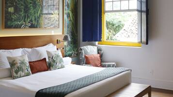 Quarto com uma cama grande, um banco, uma janela com persianas amarelas e obras de arte com tema tropical.