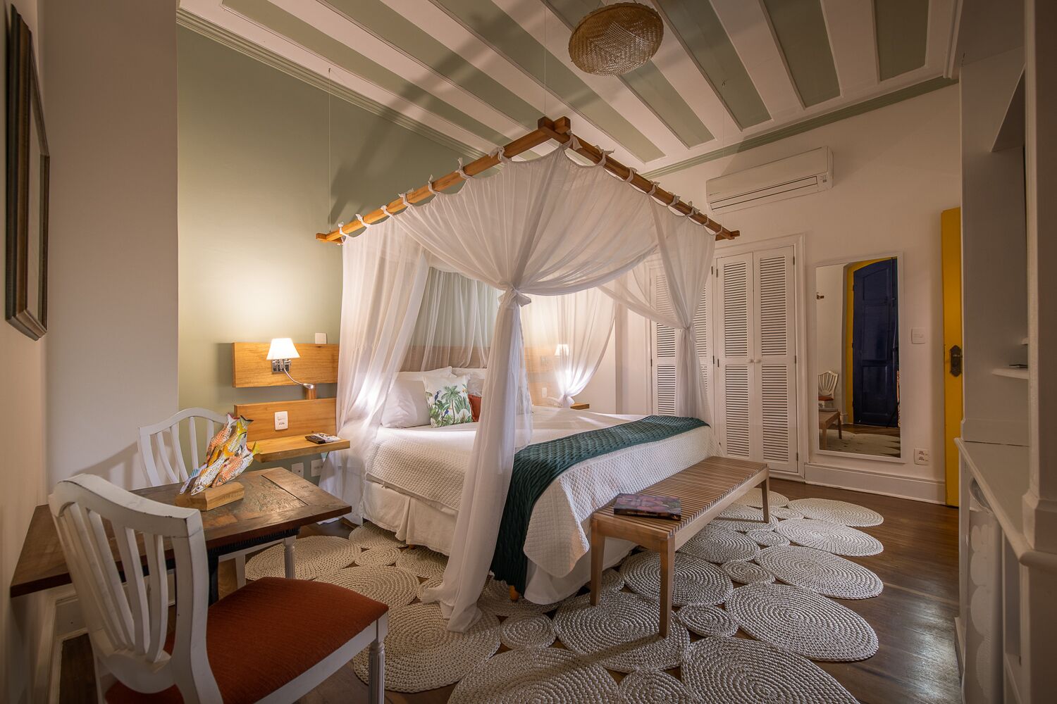 Quarto com cama de dossel, mesa de cabeceira com abajur, cadeira e tapete redondo de fibra.