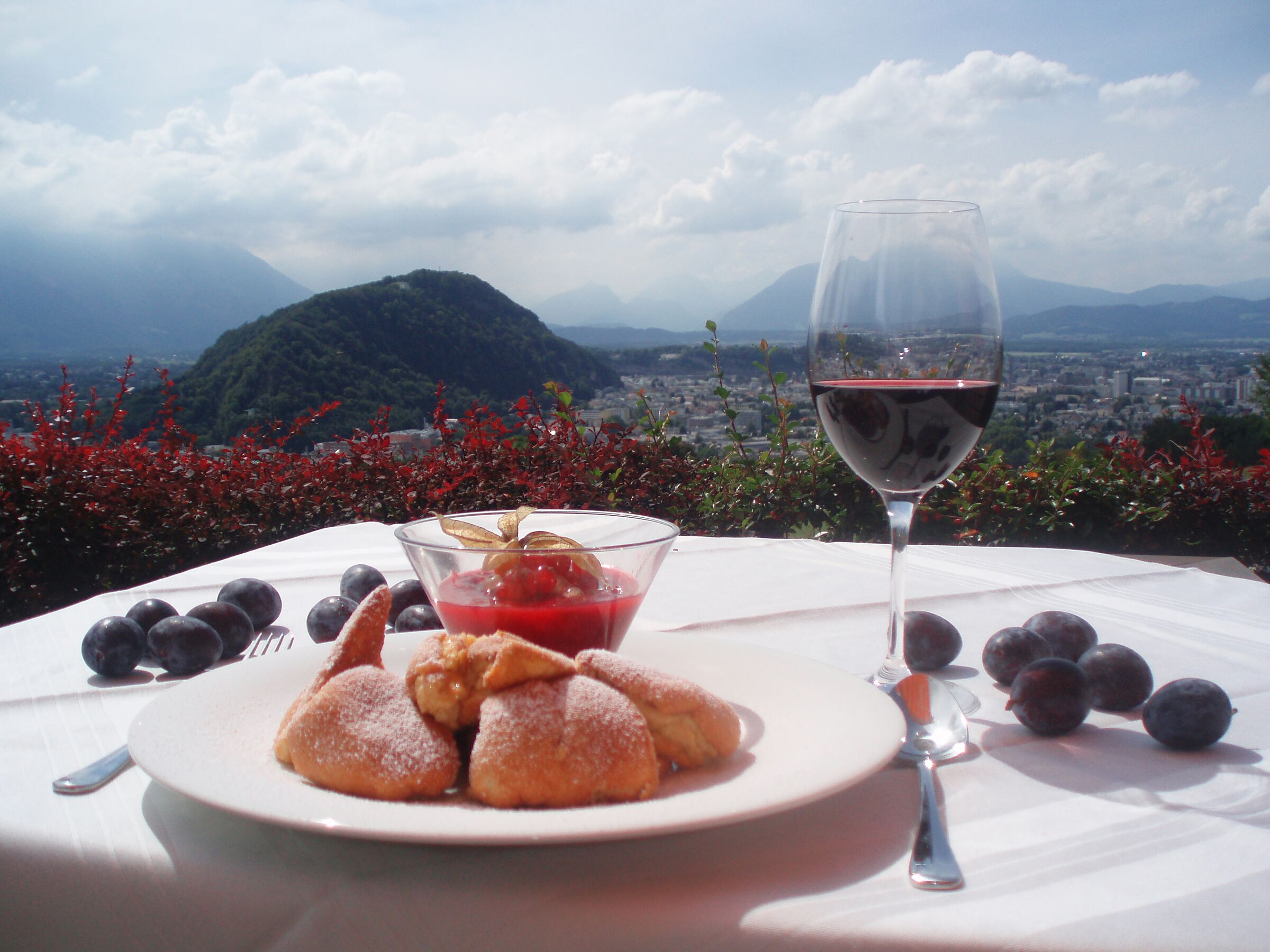 Uma mesa com um prato de doces, um copo de vinho tinto e uma taça de frutas sobre uma toalha, com vista para uma paisagem urbana e montanhas ao fundo.