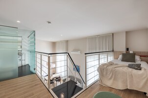 Quarto de hotel moderno com divisória de vidro, escada, cama com manta bege e vista para a cidade.