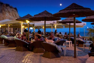 Um restaurante à beira-mar com mesas ao ar livre, guarda-sóis de palha e vista para o oceano ao entardecer.