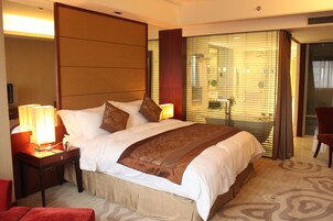 Quarto de hotel com cama grande, abajures, uma cadeira vermelha e banheiro com chuveiro de vidro.
