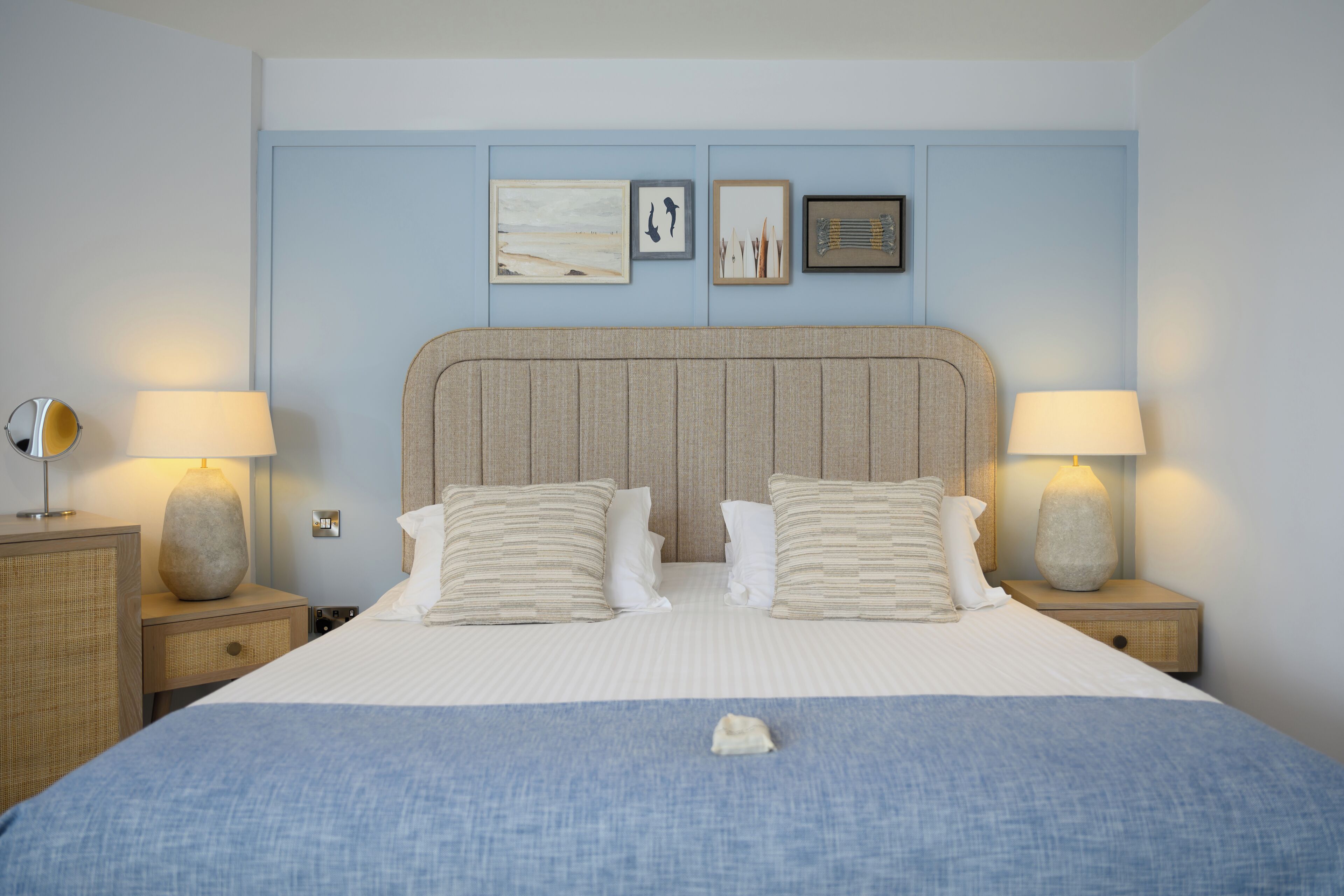 Uma cama de casal bem arrumada com colcha azul, duas mesas de cabeceira com abajures e uma obra de arte na parede acima da cama.