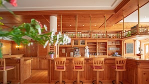 Uma área de bar com balcão de madeira, banquetes altos, uma máquina de café e prateleiras com copos.