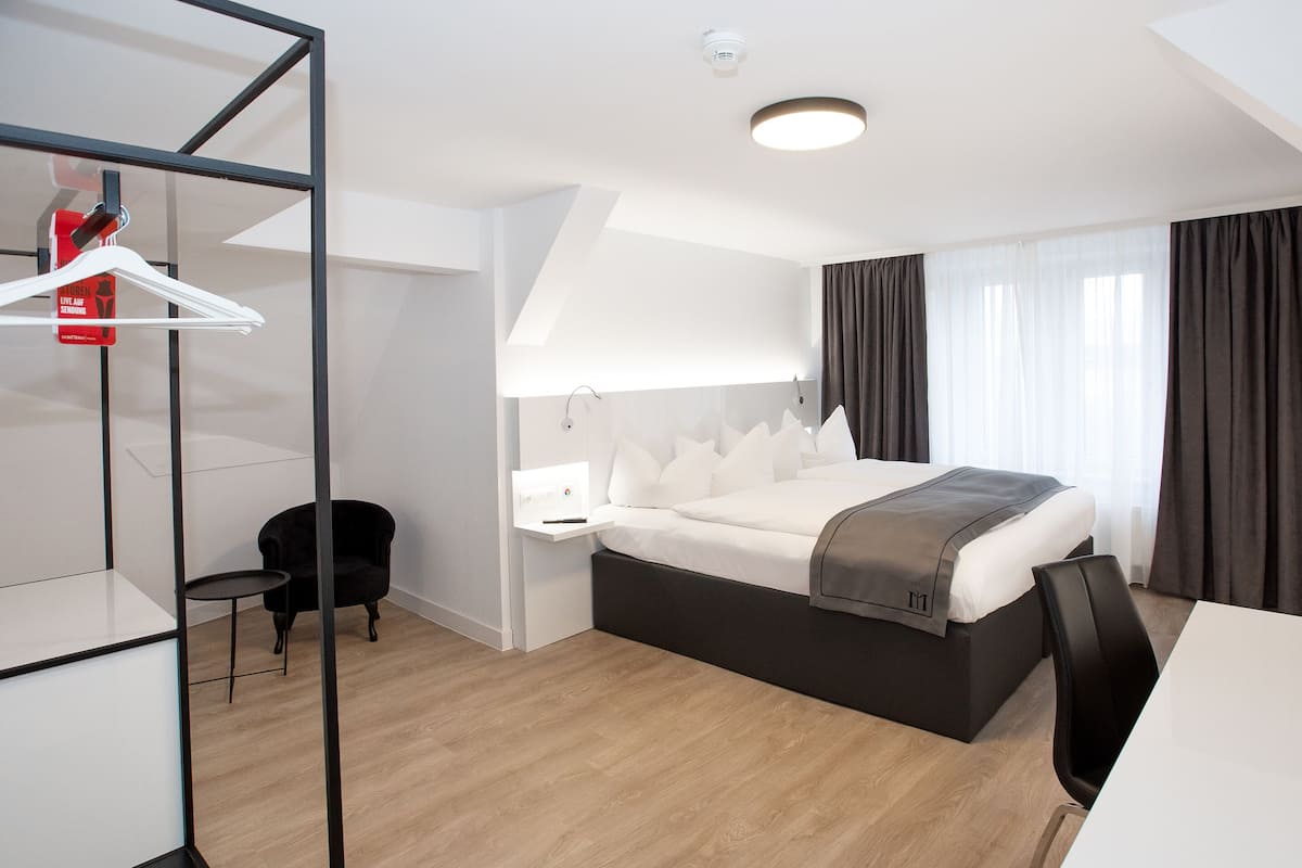 Quarto de hotel moderno com uma cama grande, uma escrivaninha, uma cadeira, um guarda-roupa e um cabideiro.