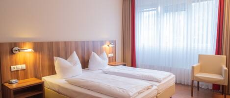 Quarto de hotel com cama de casal, mesas de cabeceira, uma cadeira e uma janela grande com cortinas.