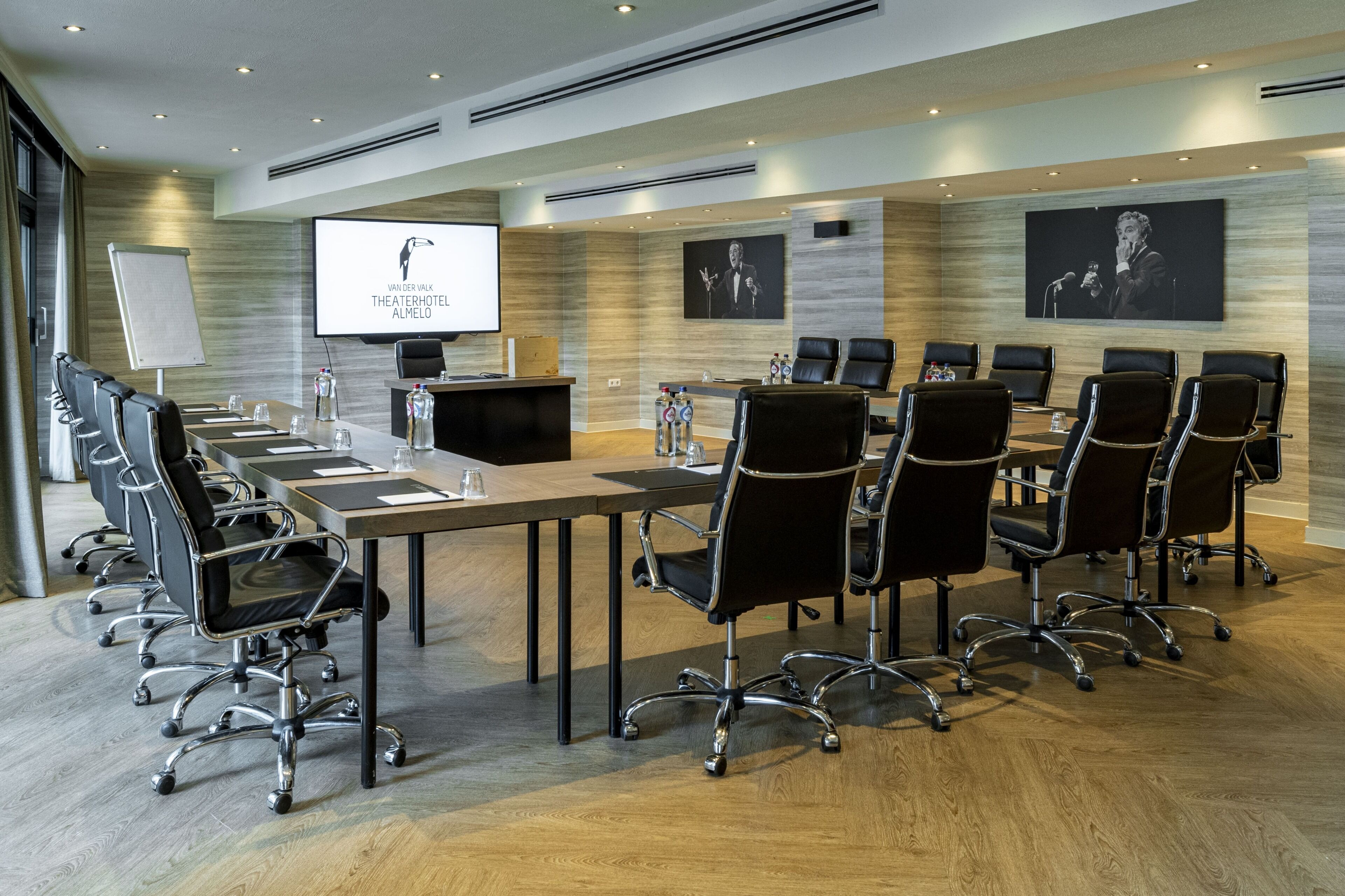 Uma sala de conferência com uma mesa comprida, cadeiras pretas, um púlpito e uma tela exibindo 'VAN DER VALK THEATERHOTEL ALMELO'.