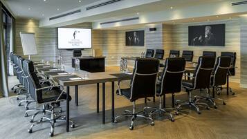Uma sala de conferência com uma mesa comprida, cadeiras pretas, um púlpito e uma tela exibindo 'VAN DER VALK THEATERHOTEL ALMELO'.
