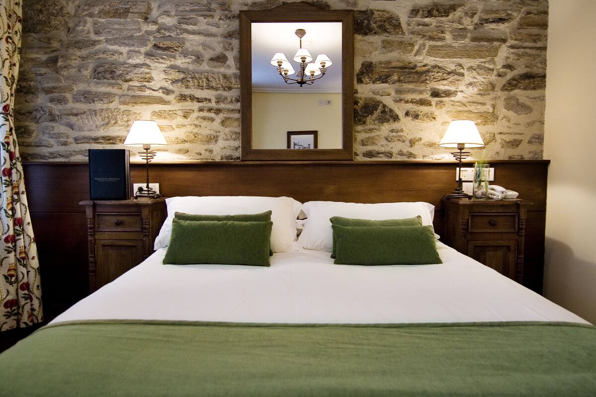 Cama bem arrumada com almofadas verdes, cabeceira de madeira e parede de pedra ao fundo. Há duas luminárias de apoio e um quadro na cabeceira.