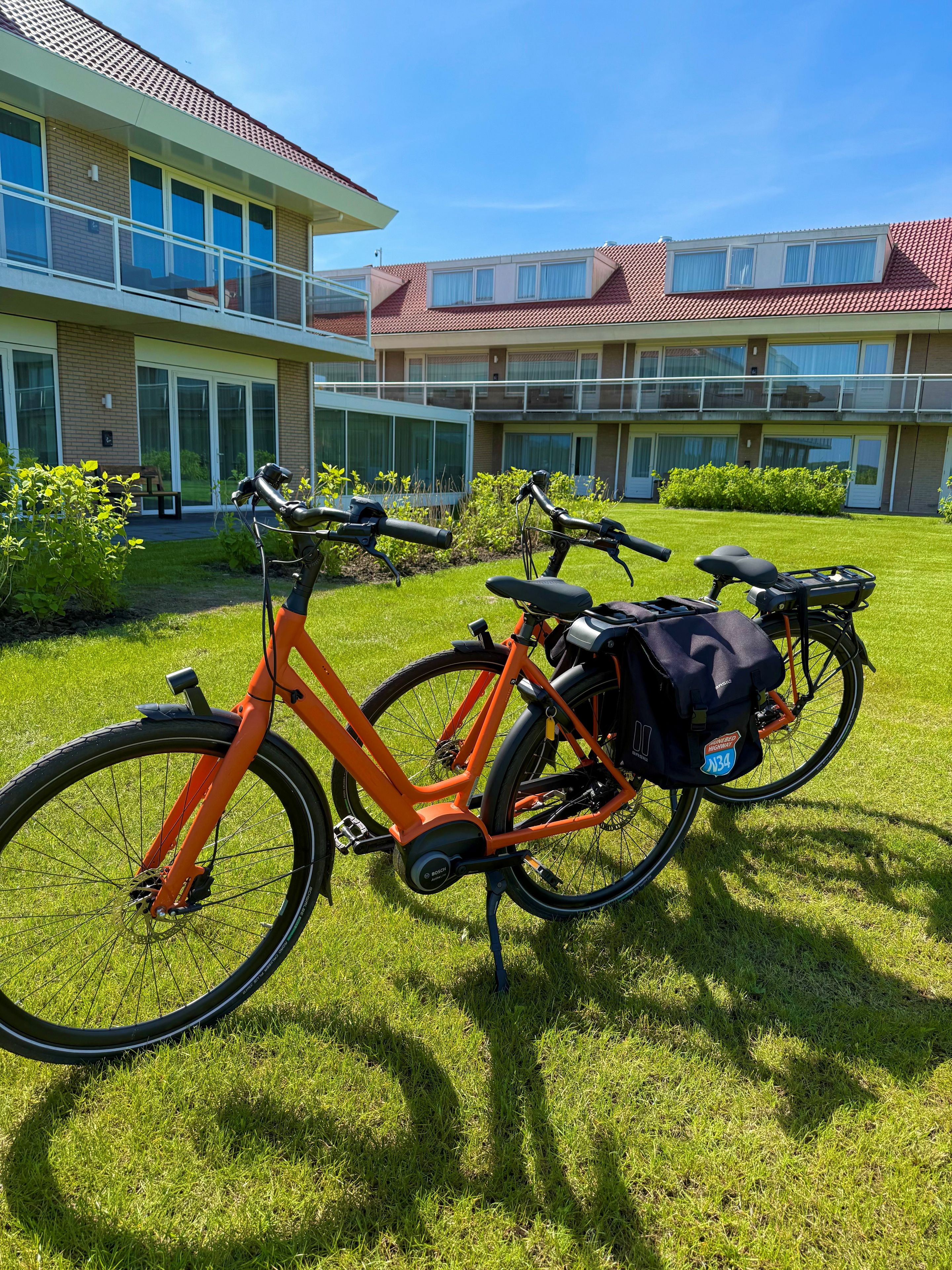 Bicicleta com uma bolsa preta, estacionada em área gramada em frente a um edifício com várias janelas e sacadas.