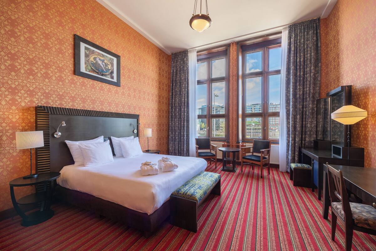Quarto de hotel com uma cama grande, uma escrivaninha, uma cadeira, uma mesinha e uma janela com cortinas.