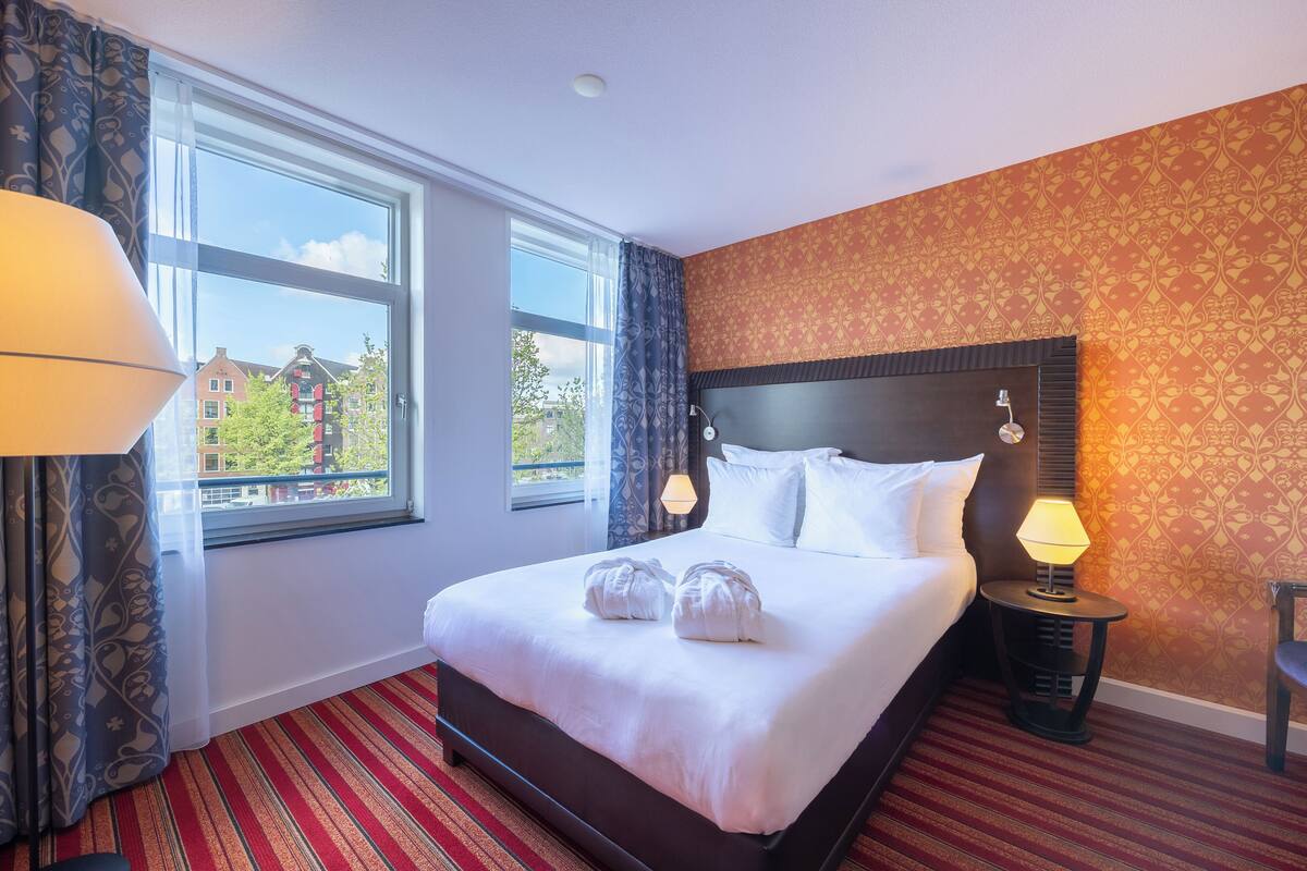 Quarto de hotel com uma cama grande, duas luminárias de cabeceira, uma janela com cortinas e parede com padrão.