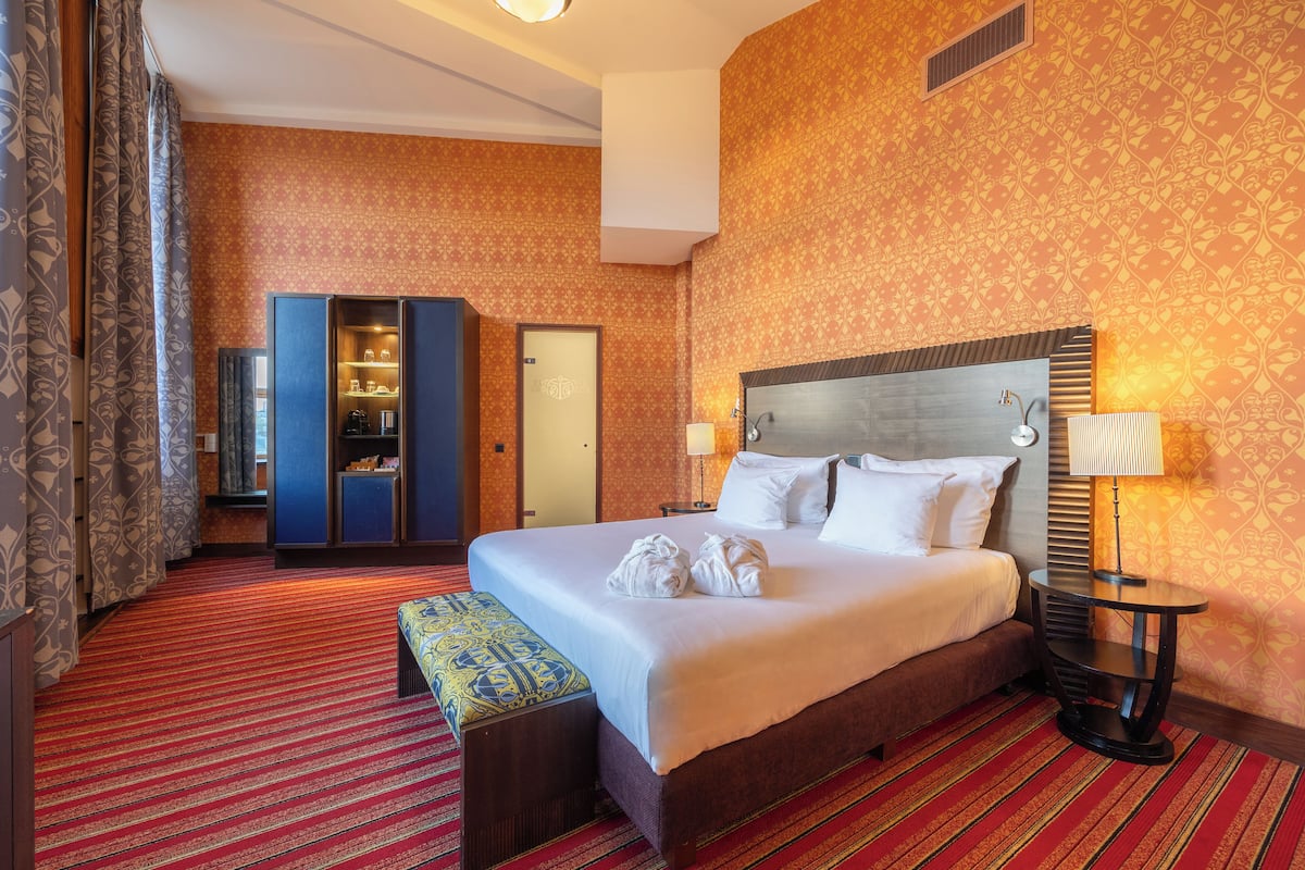Quarto de hotel com uma cama grande, luminárias de cabeceira, um guarda-roupa e um banco.