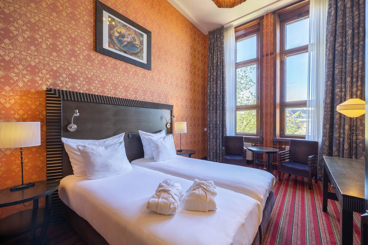 Quarto de hotel com uma cama grande, duas mesas de cabeceira, uma escrivaninha, uma cadeira e uma janela com cortinas.
