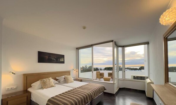 Quarto de hotel com uma janela grande, uma cama, uma mesa de cabeceira e uma cadeira perto da janela.